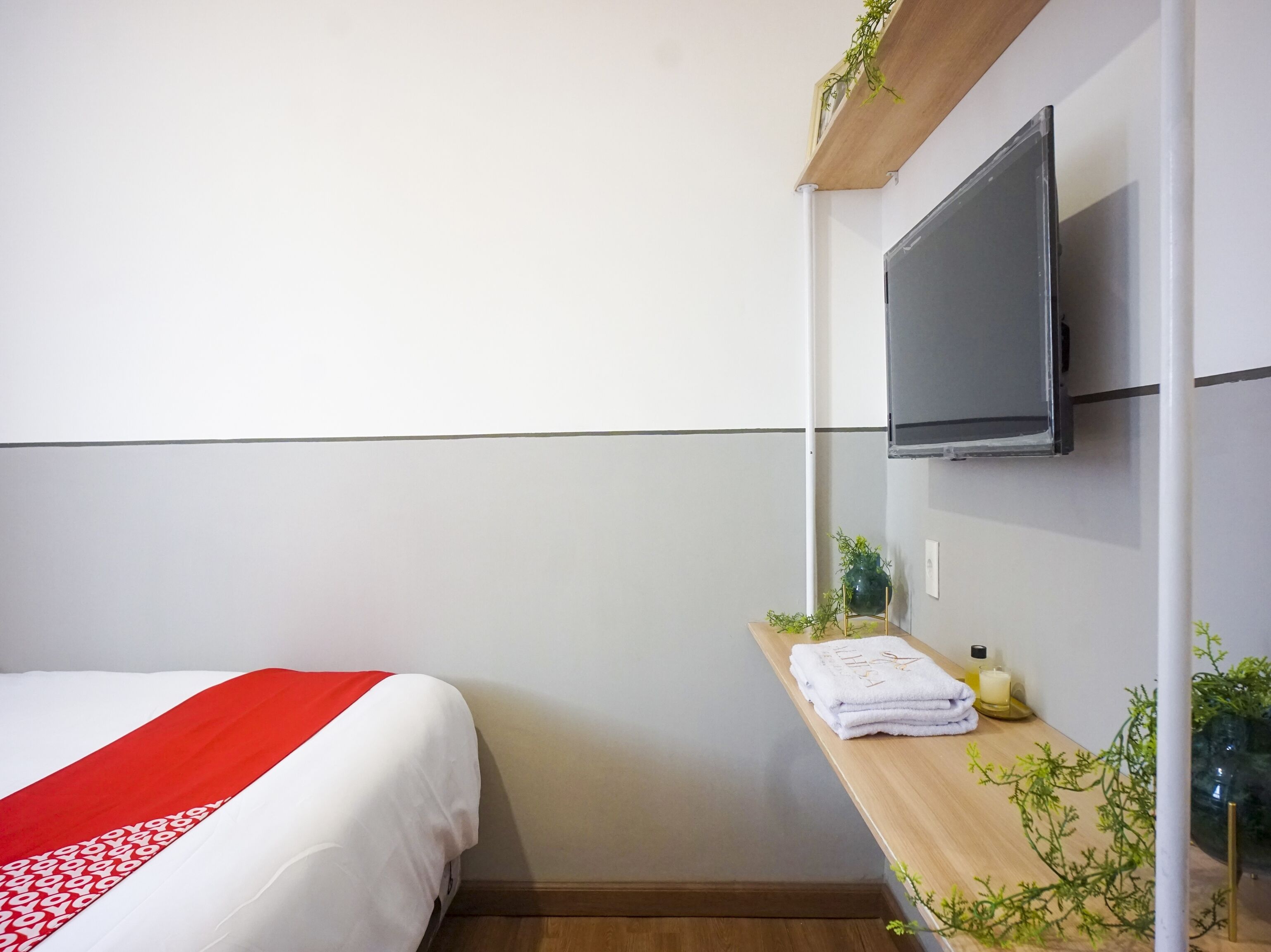 Quarto de hotel com cama, televisão fixada na parede, prateleira de madeira pequena com toalhas e uma vela, e planta em vaso.
