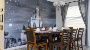 Uma sala de jantar com uma mesa posta para quatro pessoas, rodeada por cadeiras de madeira com assentos acolchoados. Um candelabro pendurado sobre a mesa, e um mural grande retrata um castelo sob um céu estrelado.