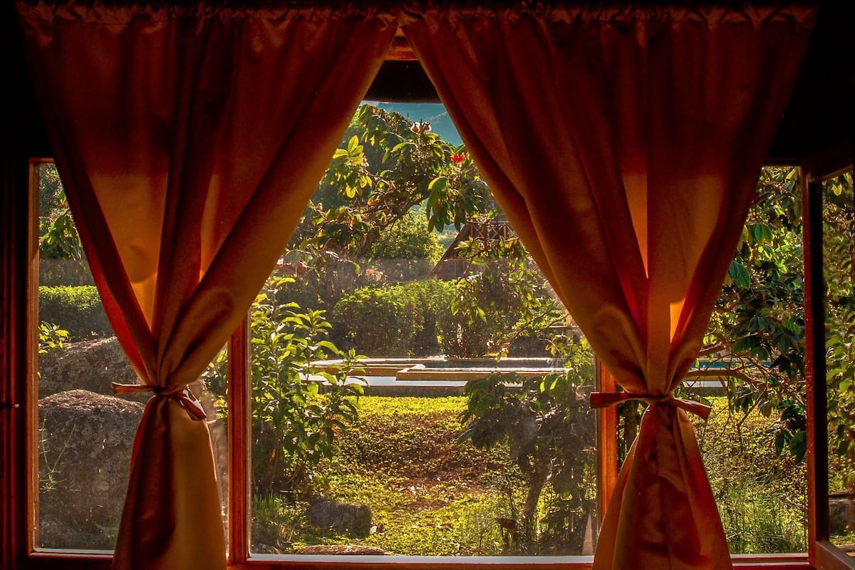 Janela com cortinas vermelhas amarradas, mostrando vista de um jardim com árvores e uma estrutura de metal.