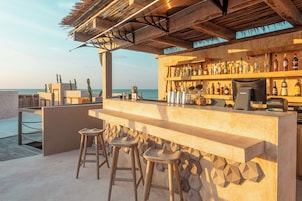 Um bar à beira-mar com balcão de madeira, banquetas e diversas garrafas.