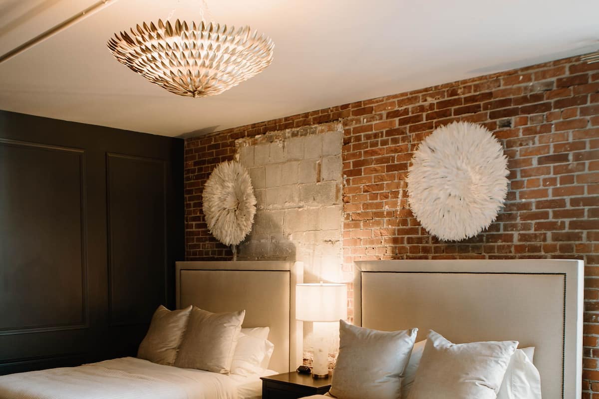 Quarto de hotel com duas camas, um tapete estampado, uma mesinha e um objeto decorativo na parede.