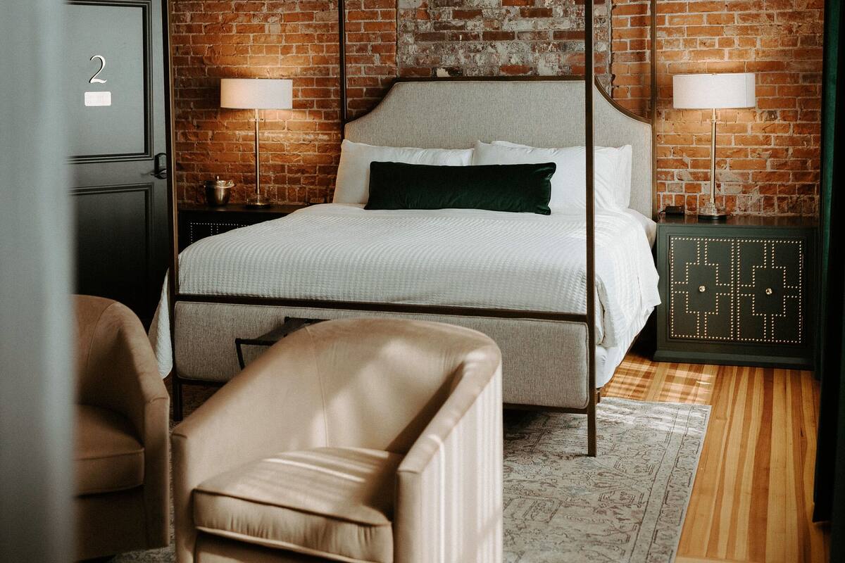 Quarto com cama de dossel, duas luminárias de cabeceira, piso de madeira e parede de tijolos.