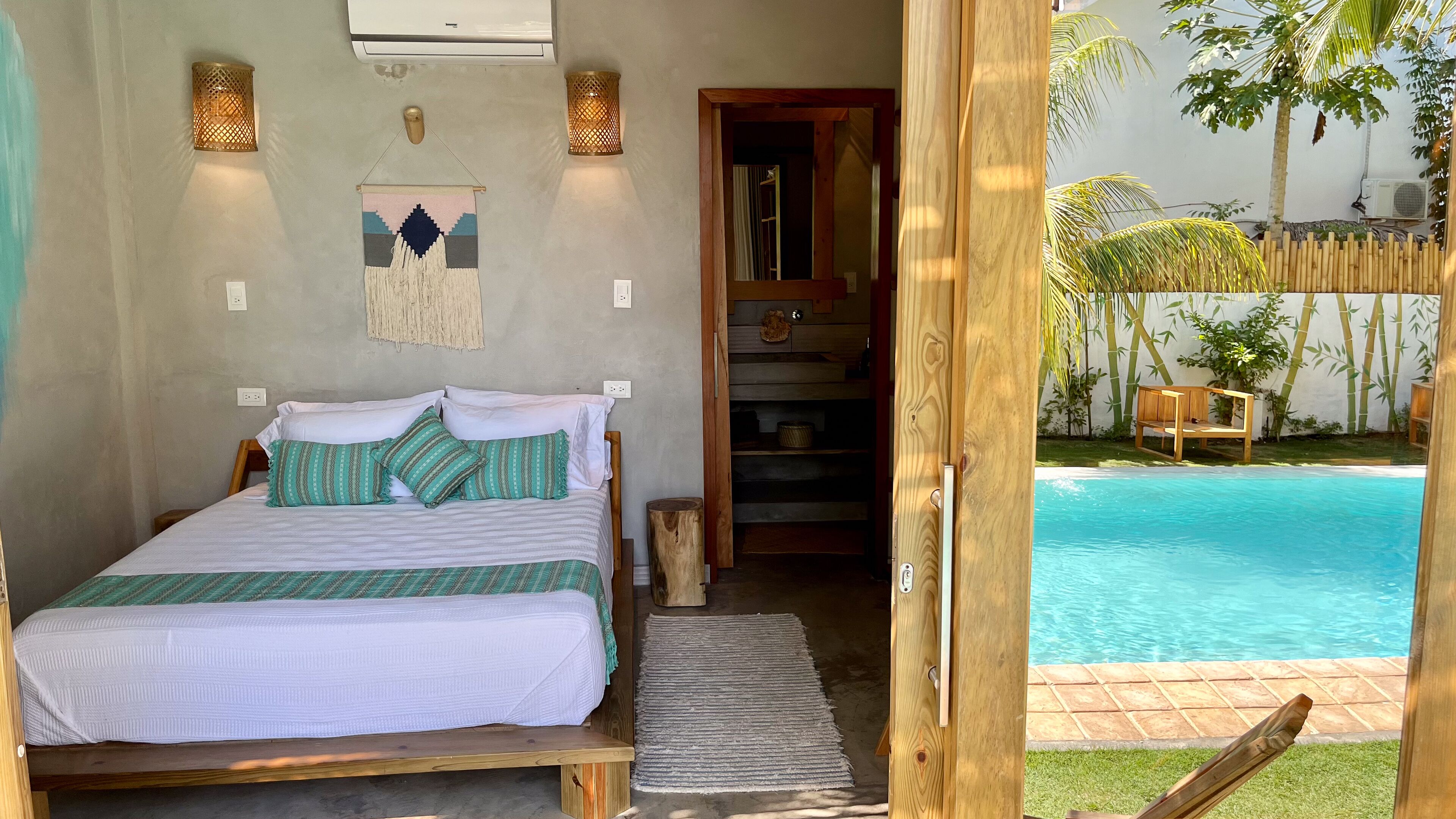 Quarto com cama, almofadas, mesa de cabeceira, luminária na parede e vista para uma piscina.