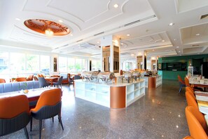 Um amplo espaço de refeições de hotel, com diversas opções de assento, um buffet central e vista para o exterior através de grandes janelas.