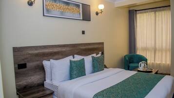 Quarto de hotel com cabeceira de madeira, cama com roupa de cama branca e verde, duas luminárias fixadas na parede, uma obra de arte emoldurada, uma poltrona verde e uma janela com cortinas.