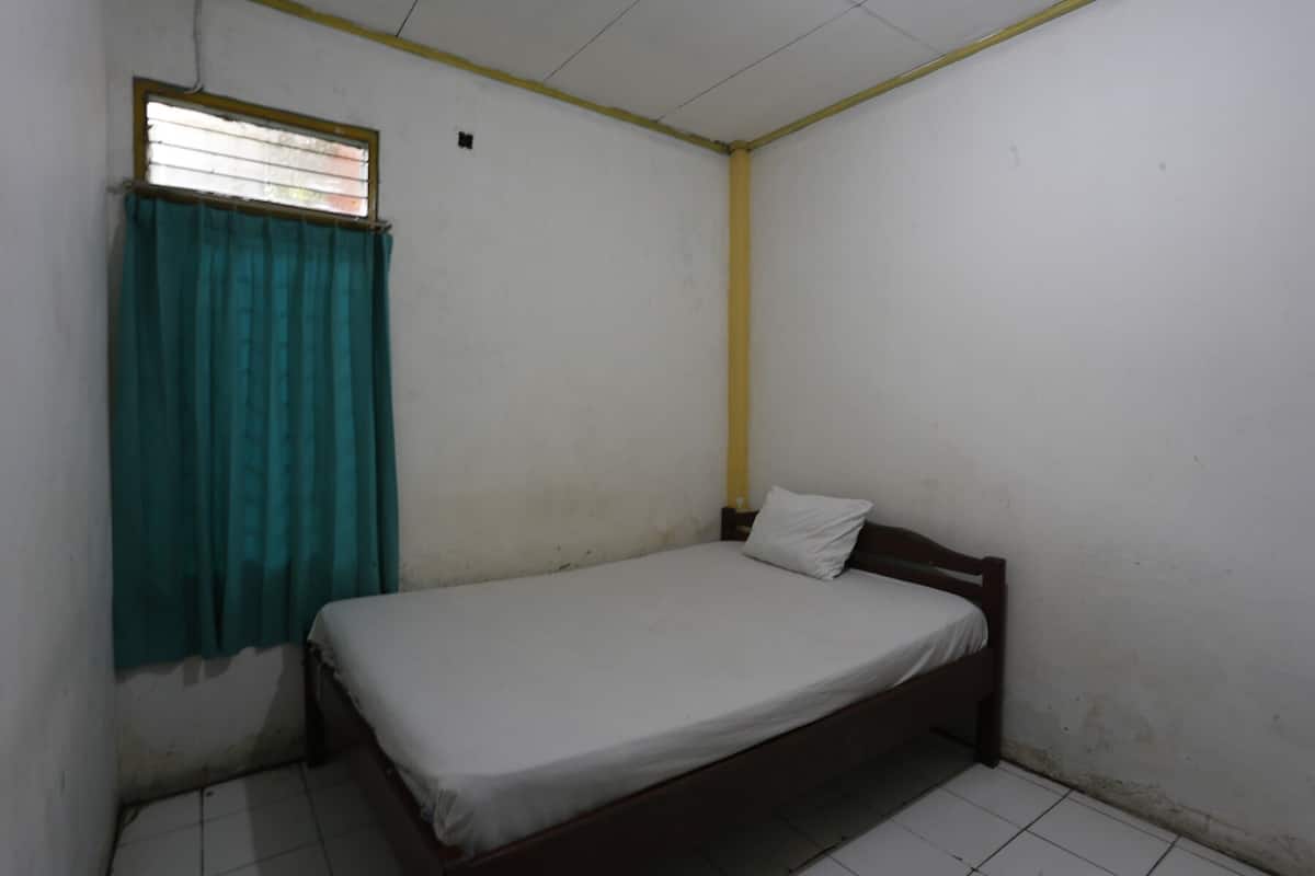 Quarto pequeno com uma cama de solteiro, janela coberta por uma cortina, parede branca e um canto pintado de amarelo.