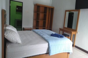 Uma cama bem arrumada com colcha branca e azul, uma mesa de cabeceira de madeira, um espelho e uma porta verde ao fundo.
