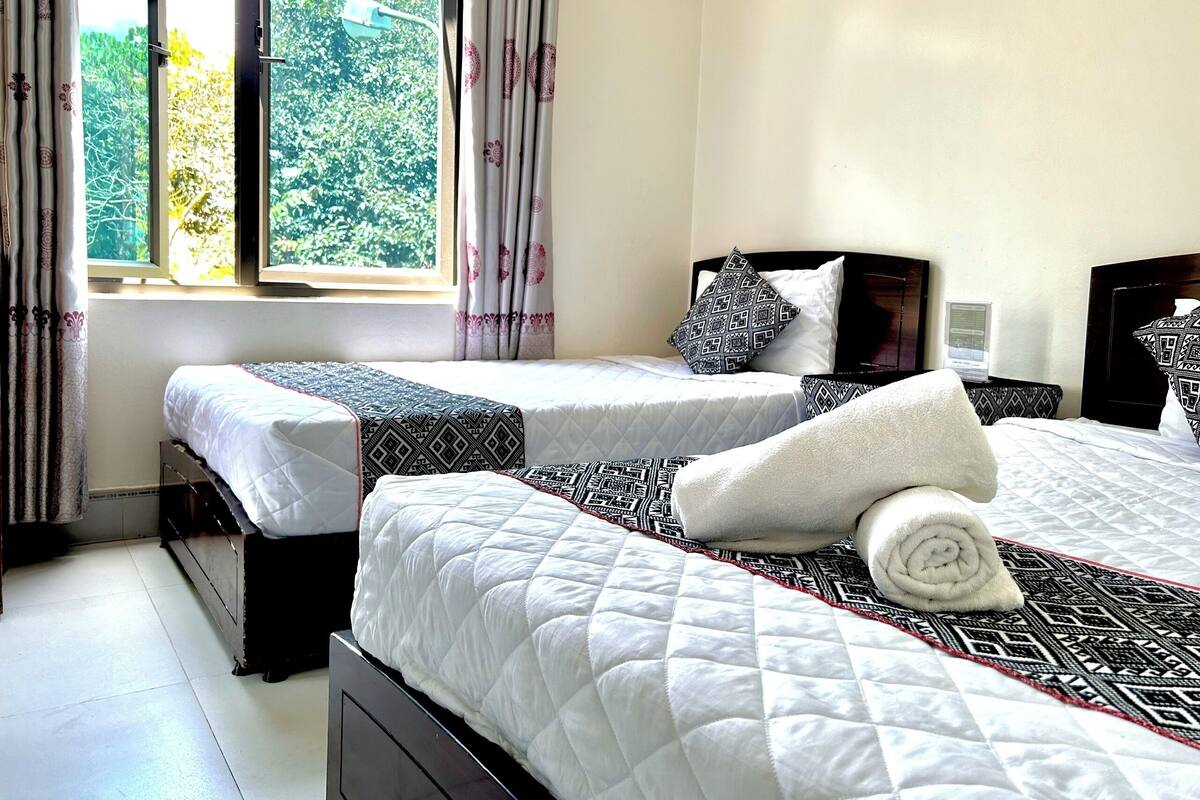 Quarto de hotel com duas camas, uma janela com cortinas e vista para áreas verdes.