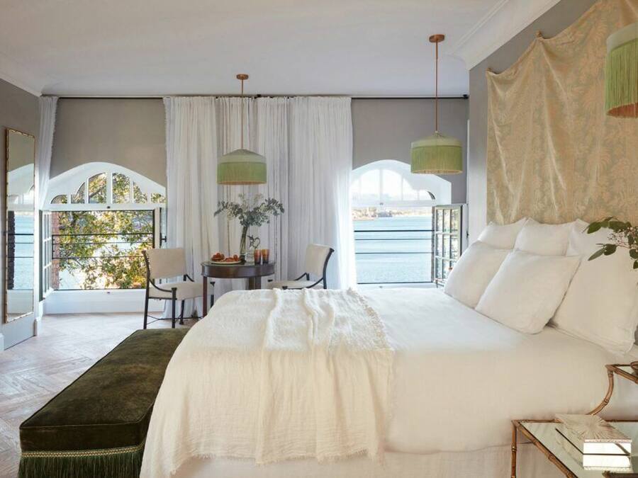 Quarto espaçoso com uma cama grande, um banquinho e vista para a água através de janelas amplas.