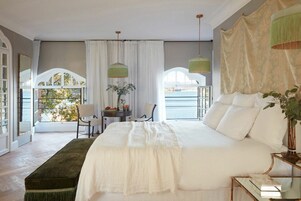 Quarto espaçoso com uma cama grande, um banquinho e vista para a água através de janelas amplas.