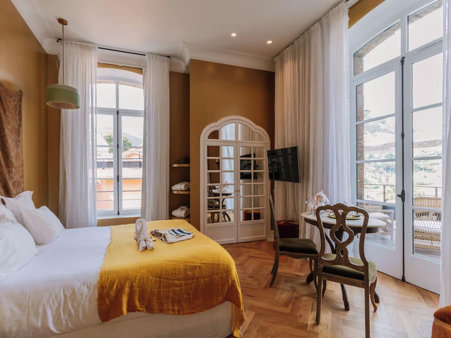 Quarto de hotel com cama, televisão, área de estar com mesa e cadeiras, e vista para o exterior.