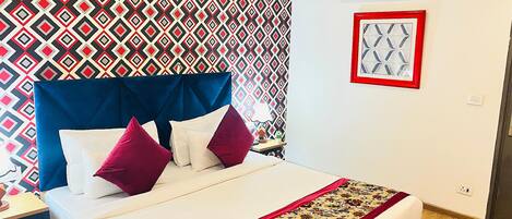 Cama bem arrumada com cabeceira azul, roupa de cama branca e manta estampada. Uma parede decorada com padrão e um quadro complementam o ambiente.