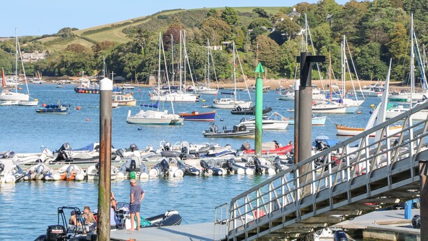 Uma marina com vários barcos atracados, uma ponte para pedestres e uma paisagem acidentada ao fundo.
