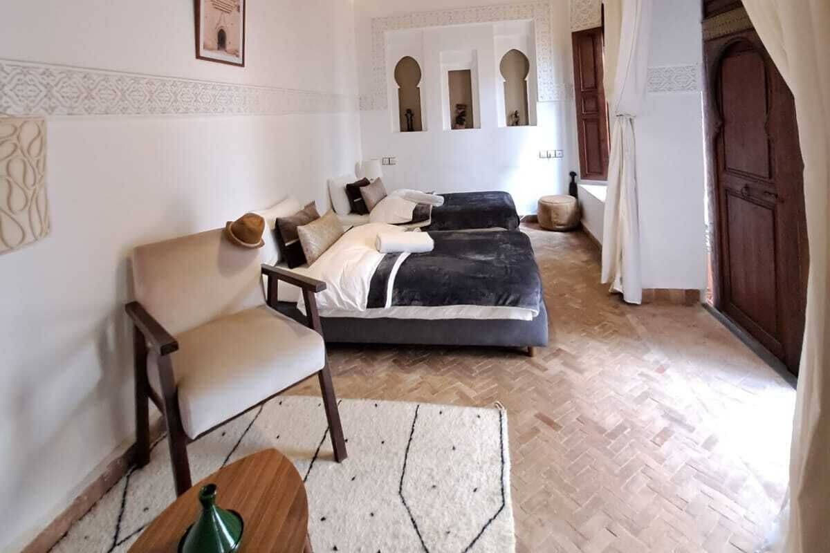 Quarto com duas camas, uma cadeira, uma mesinha e uma janela grande com cortinas.