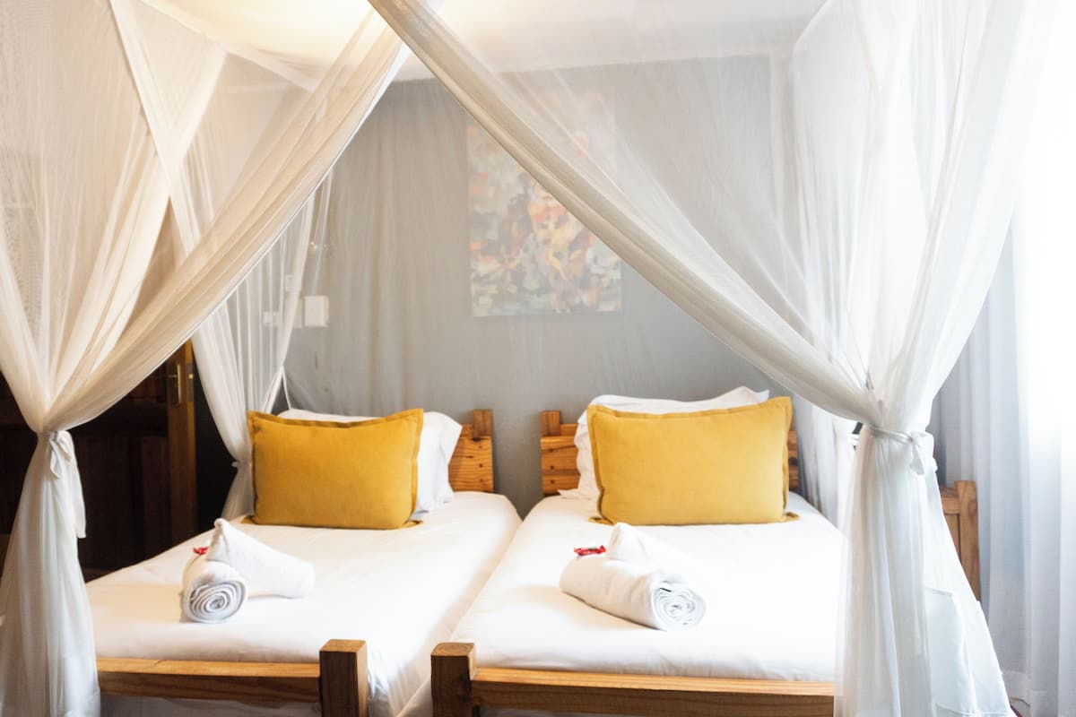 Quarto duplo com duas camas, cada uma com roupa de cama branca e almofadas amarelas, sob um dossel.