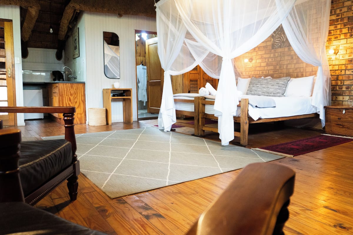 Quarto acolhedor com piso de madeira, cama com dossel, uma cadeira, uma pequena mesa e vista para uma cozinha e banheiro por uma porta aberta.
