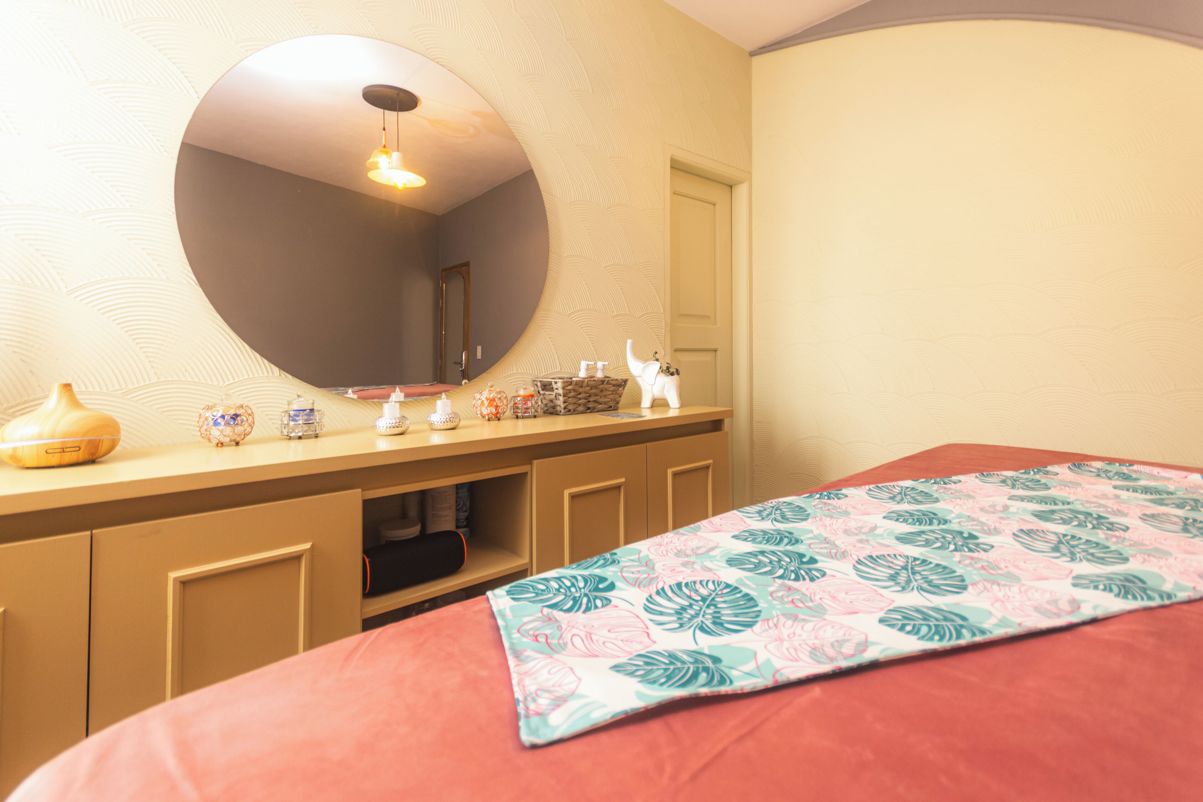 Quarto com cama, espelho redondo, penteadeira com artigos de higiene e uma pequena estante com objetos decorativos.