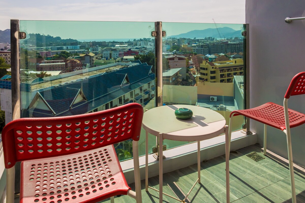 Varanda com duas cadeiras vermelhas e uma mesa pequena, com vista para uma paisagem urbana com edifícios e uma montanha ao longe.