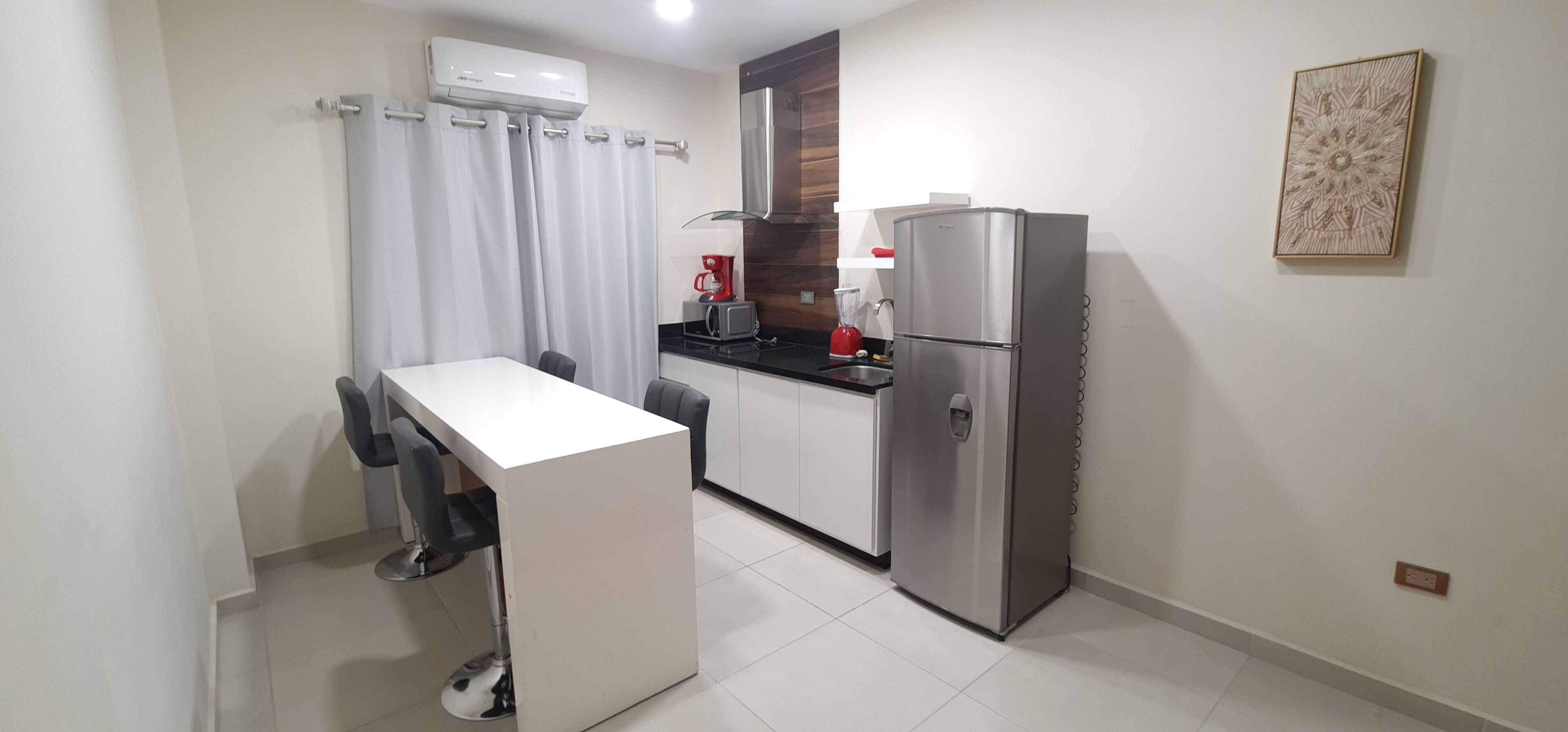Uma cozinha compacta com bancada branca, geladeira de aço inoxidável, cafeteira e ar-condicionado fixado na parede.