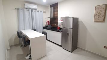 Uma cozinha compacta com bancada branca, geladeira de aço inoxidável, cafeteira e ar-condicionado fixado na parede.