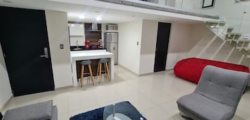 Um espaço de estar compacto com cozinha americana, área de jantar e sala de estar com um pufe vermelho e uma poltrona cinza.
