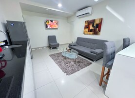 Uma sala de estar moderna com um sofá cinzento, uma mesa de centro de vidro, uma poltrona cinzenta e uma televisão fixada na parede.