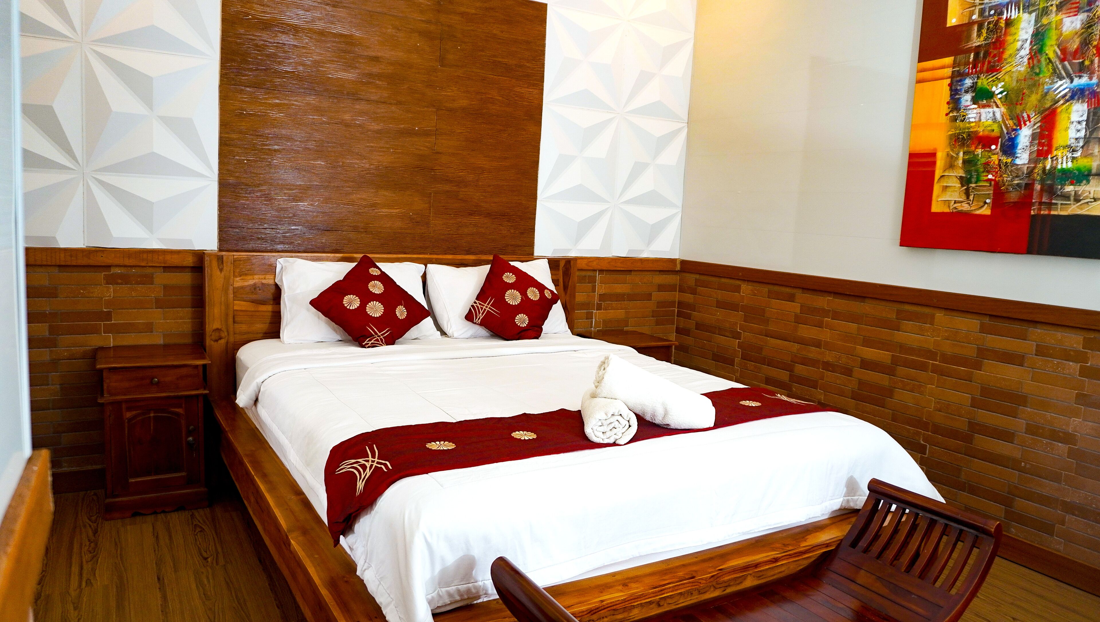 Cama bem arrumada com lençóis brancos e detalhes vermelhos, cabeceira de madeira e uma pequena mesa de cabeceira de madeira.