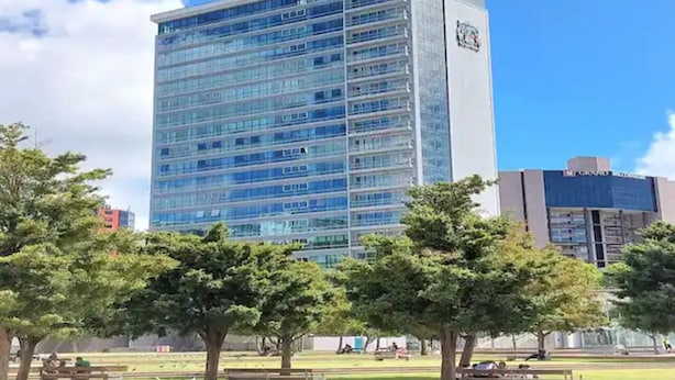 Um moderno edifício alto com fachada de vidro, rodeado por um parque com árvores e bancos.
