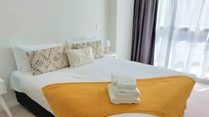 Cama bem arrumada com manta amarela e toalhas dobradas, roupa de cama branca e almofadas estampadas.
