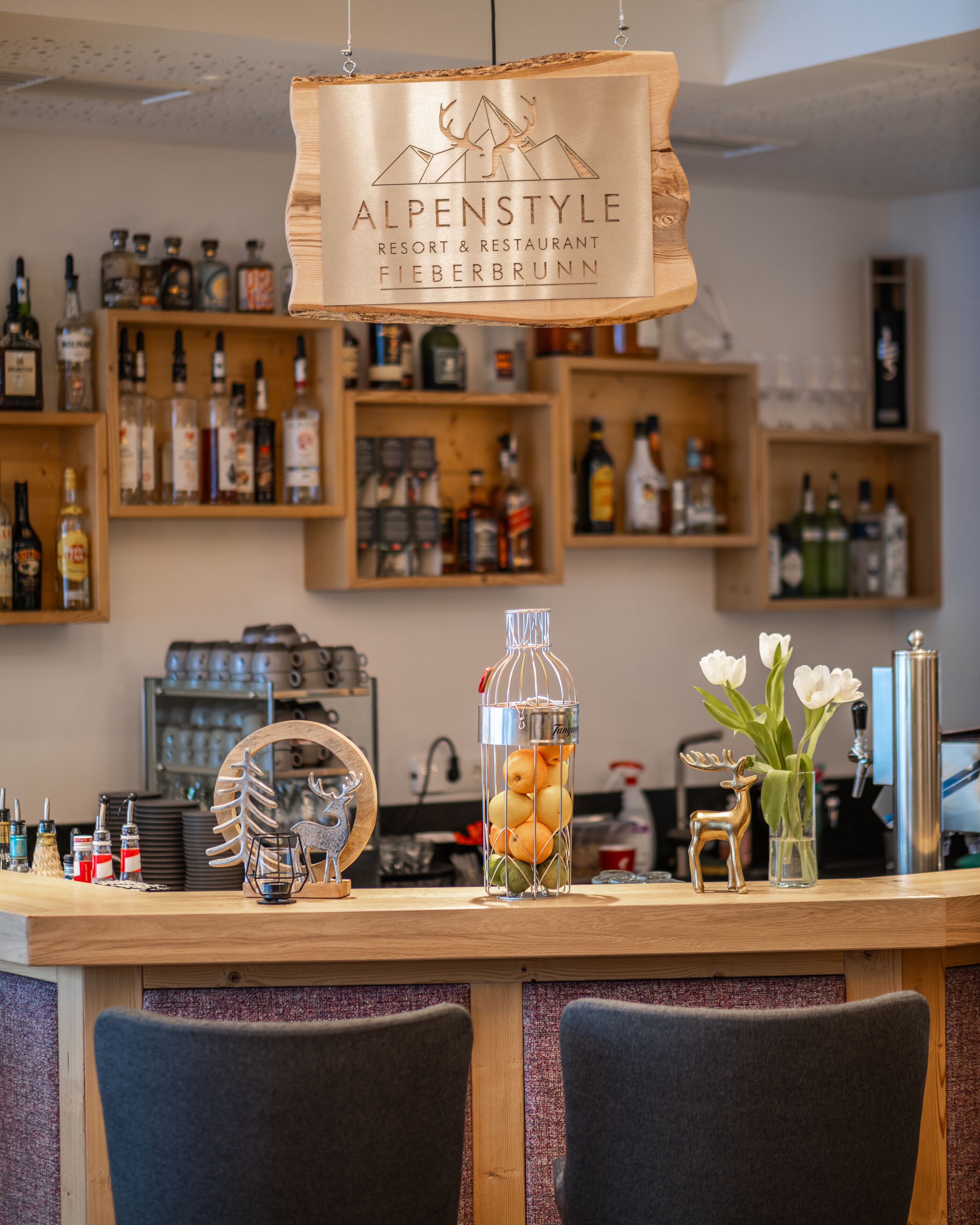 Uma área de bar com uma placa de madeira que diz "ALPENSTYLE RESORT & RESTAURANT FIEBERBRÜNN", prateleiras com garrafas de bebidas, uma cesta de frutas e uma estatueta decorativa de veado.