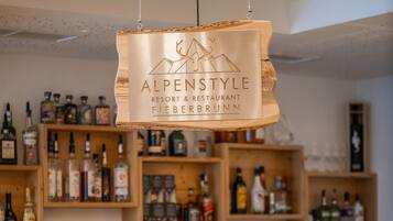 Uma área de bar com uma placa de madeira que diz "ALPENSTYLE RESORT & RESTAURANT FIEBERBRÜNN", prateleiras com garrafas de bebidas, uma cesta de frutas e uma estatueta decorativa de veado.