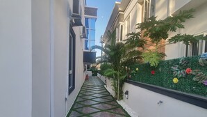Um beco estreito com chão de azulejos decorativos, uma parede verde com plantas artificiais e um edifício branco com aparelhos de ar condicionado.