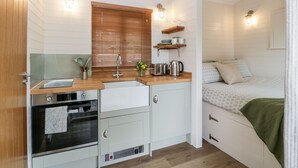 Cozinha compacta com forno embutido, pia e bancadas de madeira. Há uma janela com persianas, uma prateleira com utensílios de cozinha e, no quarto adjacente, uma cama com um cobertor verde.