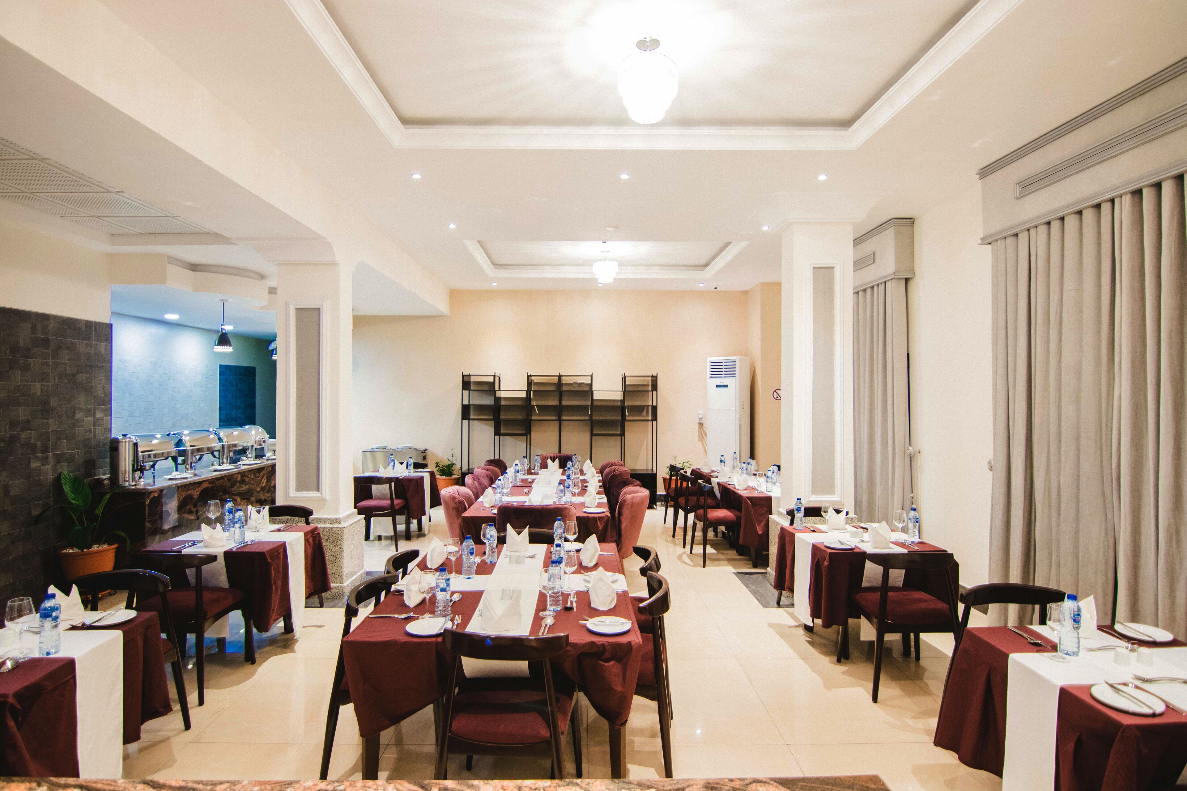 Uma área de refeições de hotel com várias mesas preparadas para o jantar, um bar e uma grande janela que proporciona iluminação natural.