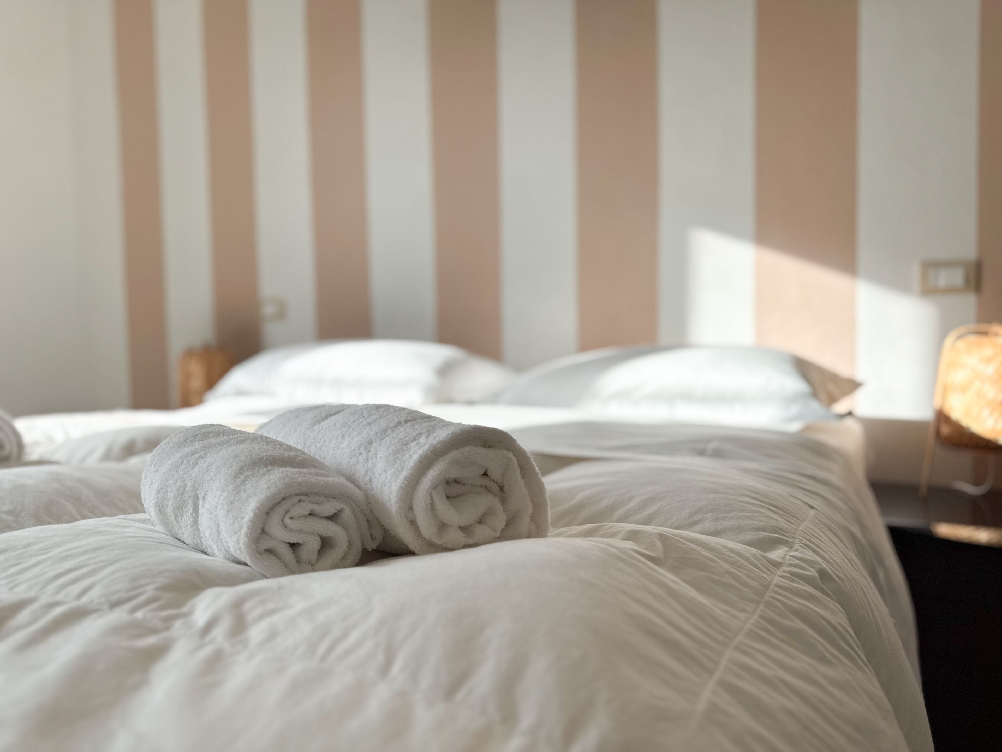 Cama bem arrumada com toalhas brancas enroladas, parede listrada e uma cadeira ao fundo.