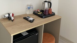 Quarto com televisão de tela plana na parede, escrivaninha de madeira com cafeteira preta, duas garrafas de água, controle remoto e um pequeno refrigerador preto.