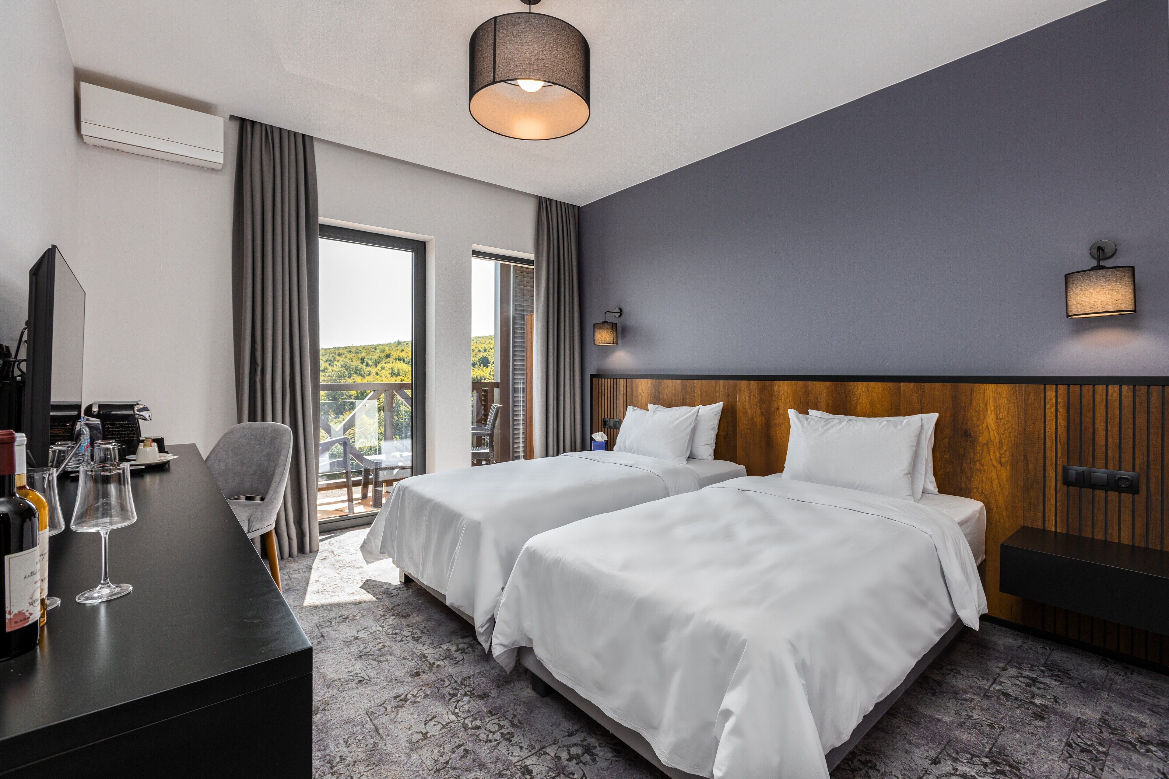 Quarto de hotel com duas camas, uma escrivaninha com computador e copos de vinho, e vista para o exterior.