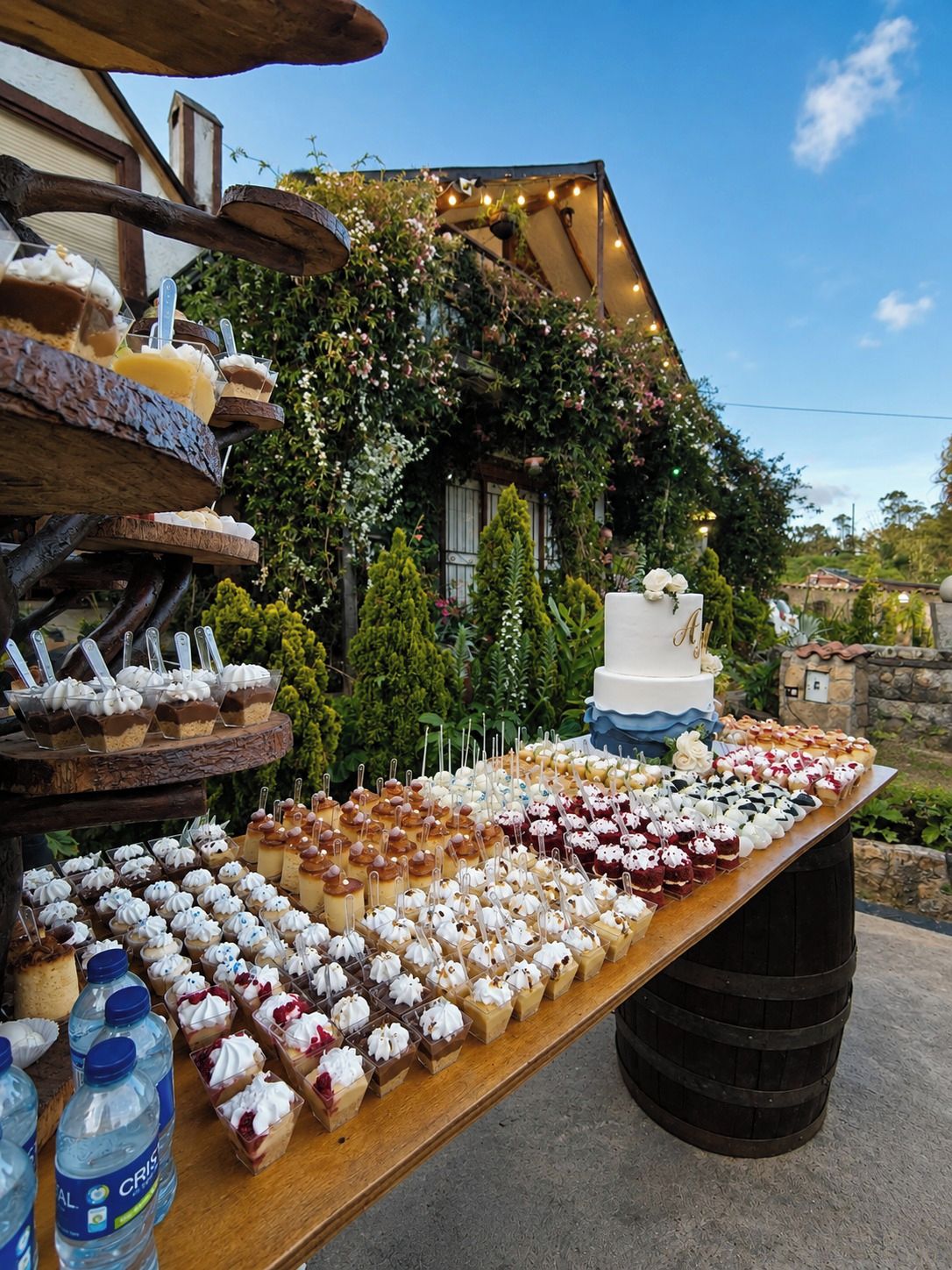 Buffet de sobremesas com diversos doces e um bolo, montado ao ar livre, com uma casa e vegetação ao fundo.