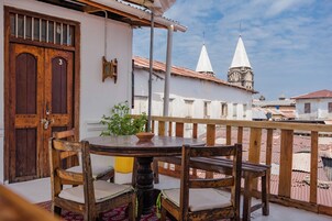 Uma varanda com mesa e cadeiras de madeira, um vaso com planta e vista para uma igreja com duas torres.
