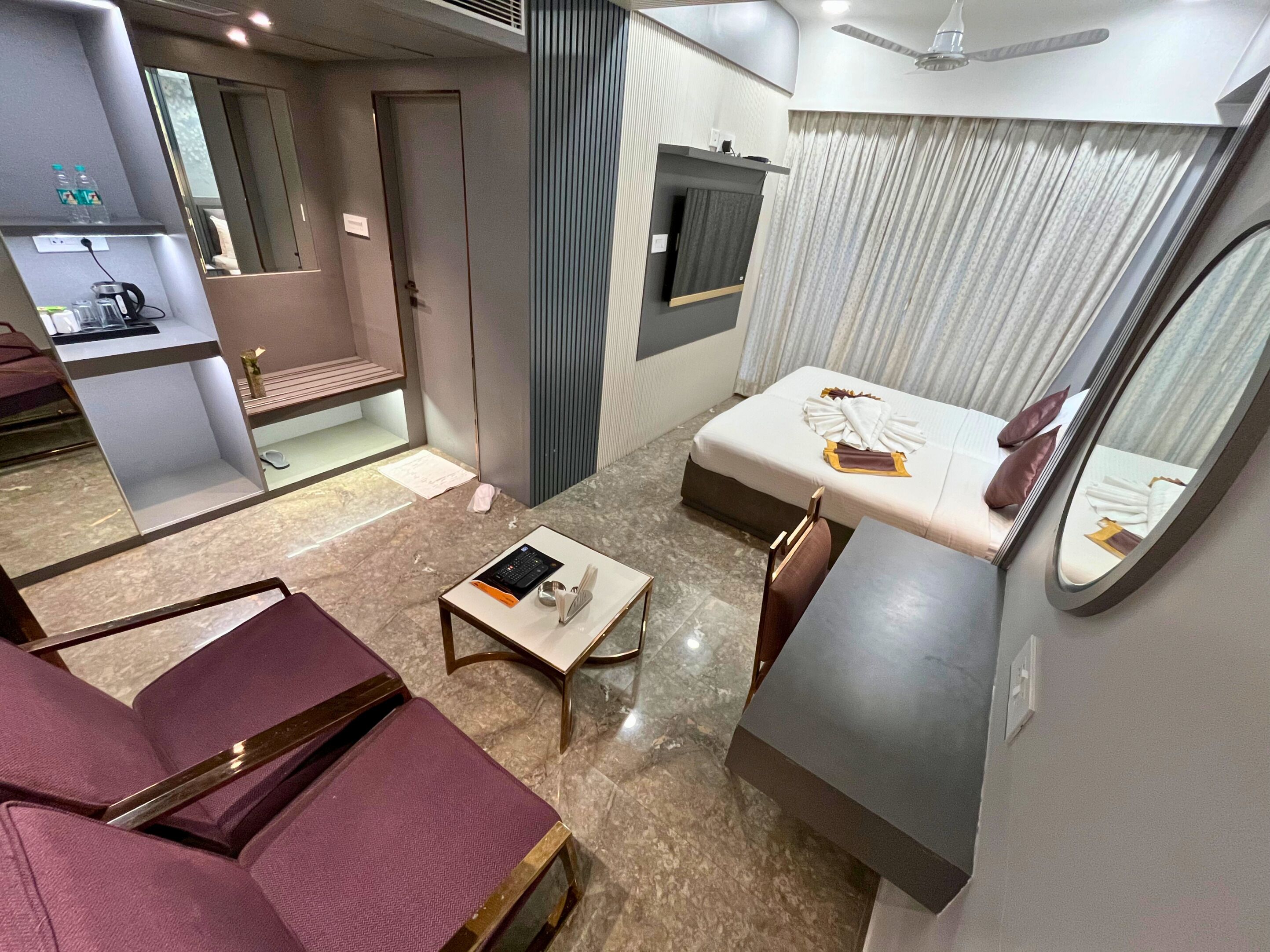 Quarto de hotel compacto com cama, uma mesinha, cadeira, escrivaninha, espelho e espaço para cozinha.