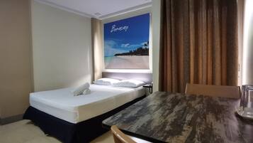 Cama de solteiro com colchão branco e saia escura, mesa de madeira e janela com vista para a praia e a palavra "Boracay".