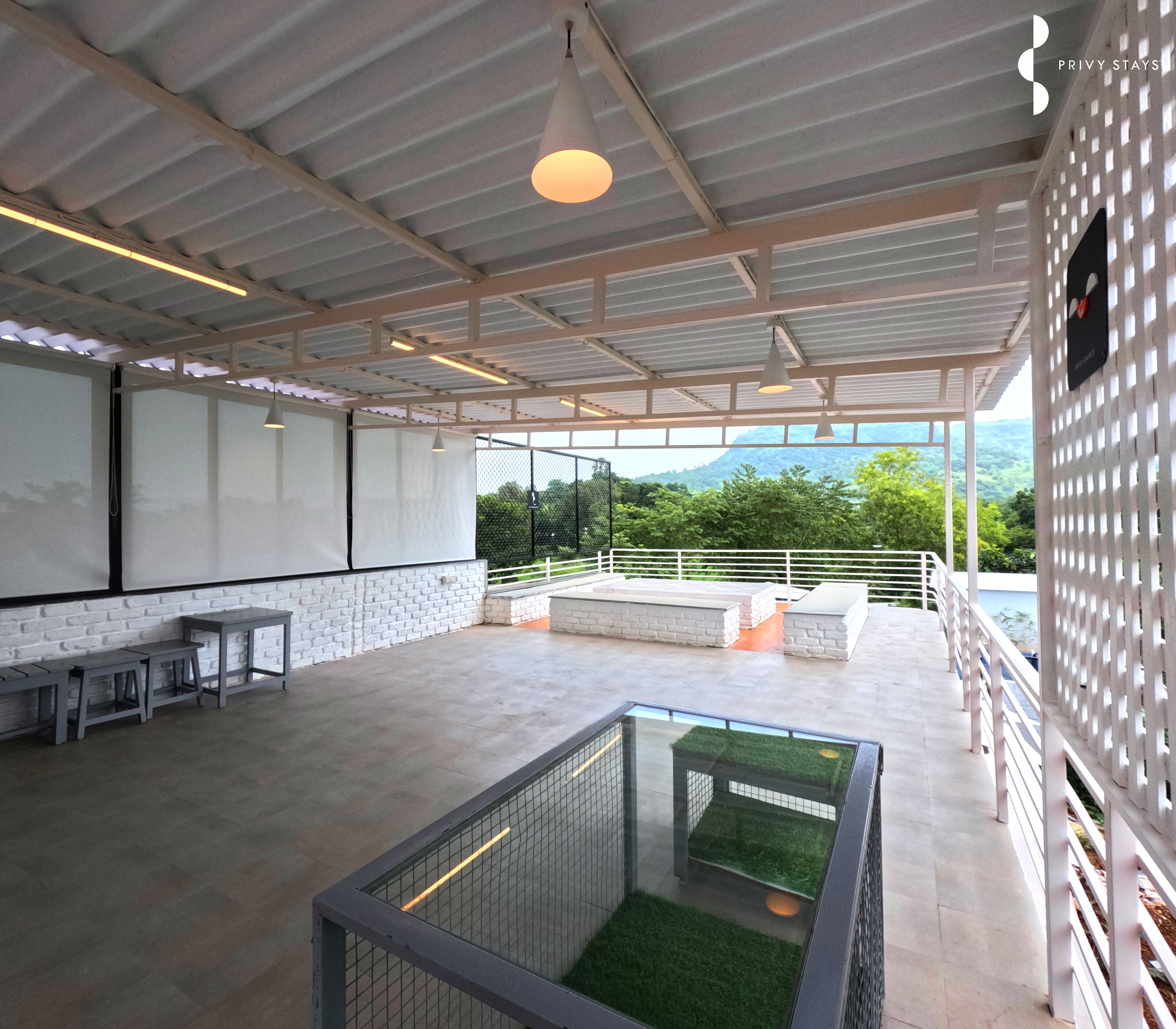 Um espaço amplo e interno com teto alto, área de estar com bancos, pequeno jardim com grama artificial e vista para uma paisagem montanhosa através de grandes janelas.