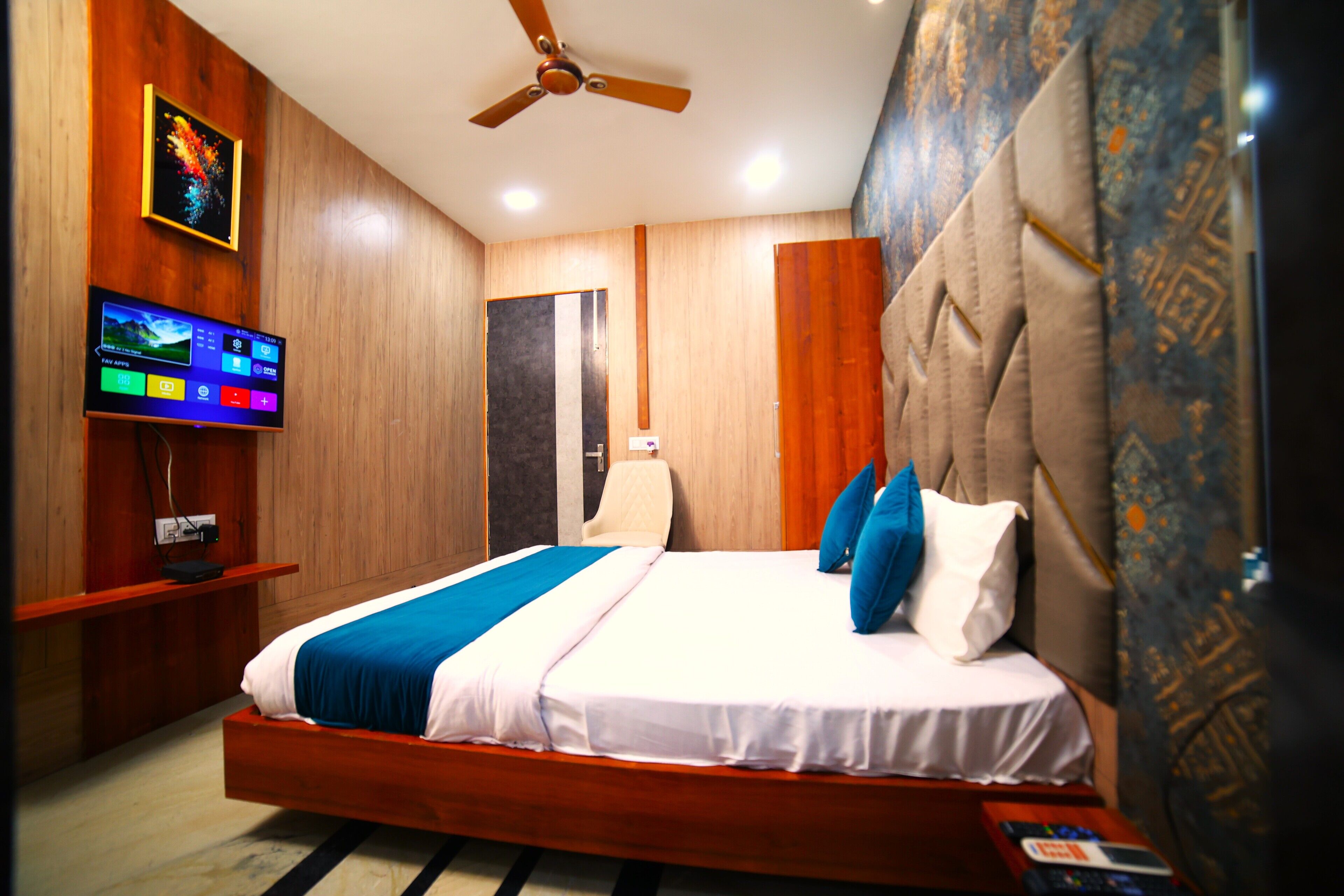 Quarto de hotel moderno com cama de madeira, TV de tela plana, ventilador de teto e uma grande janela com cortinas estampadas.
