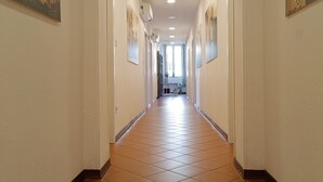 Um corredor comprido e estreito, com chão de azulejos, paredes brancas e iluminação no teto.
