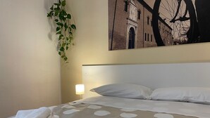 Quarto de hotel com cama, mesa de cabeceira com abajur, planta fixada na parede e um quadro grande de um edifício.