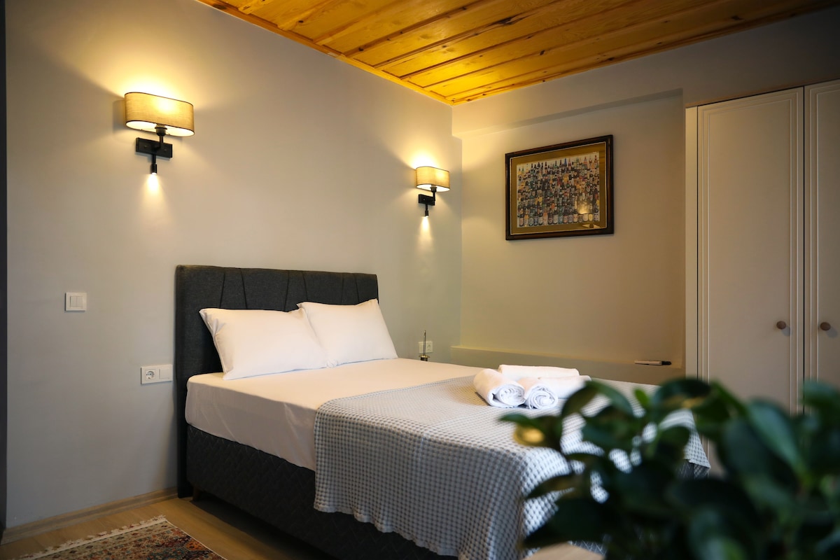 Cama bem arrumada com lençóis brancos, duas luminárias fixadas na parede, um quadro emoldurado e uma porta fechada.