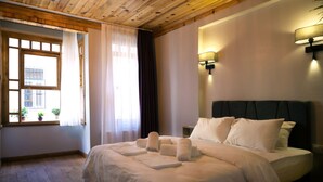 Quarto de hotel com teto de madeira, uma cama grande com roupa de cama e almofadas brancas, janela com cortinas e duas luminárias fixadas na parede.
