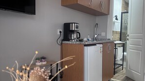A compact kitchen with a coffee maker, a small refrigerator, and wooden cabinets. There's a book titled "MUSE" on a table, and a TV mounted on the wall.