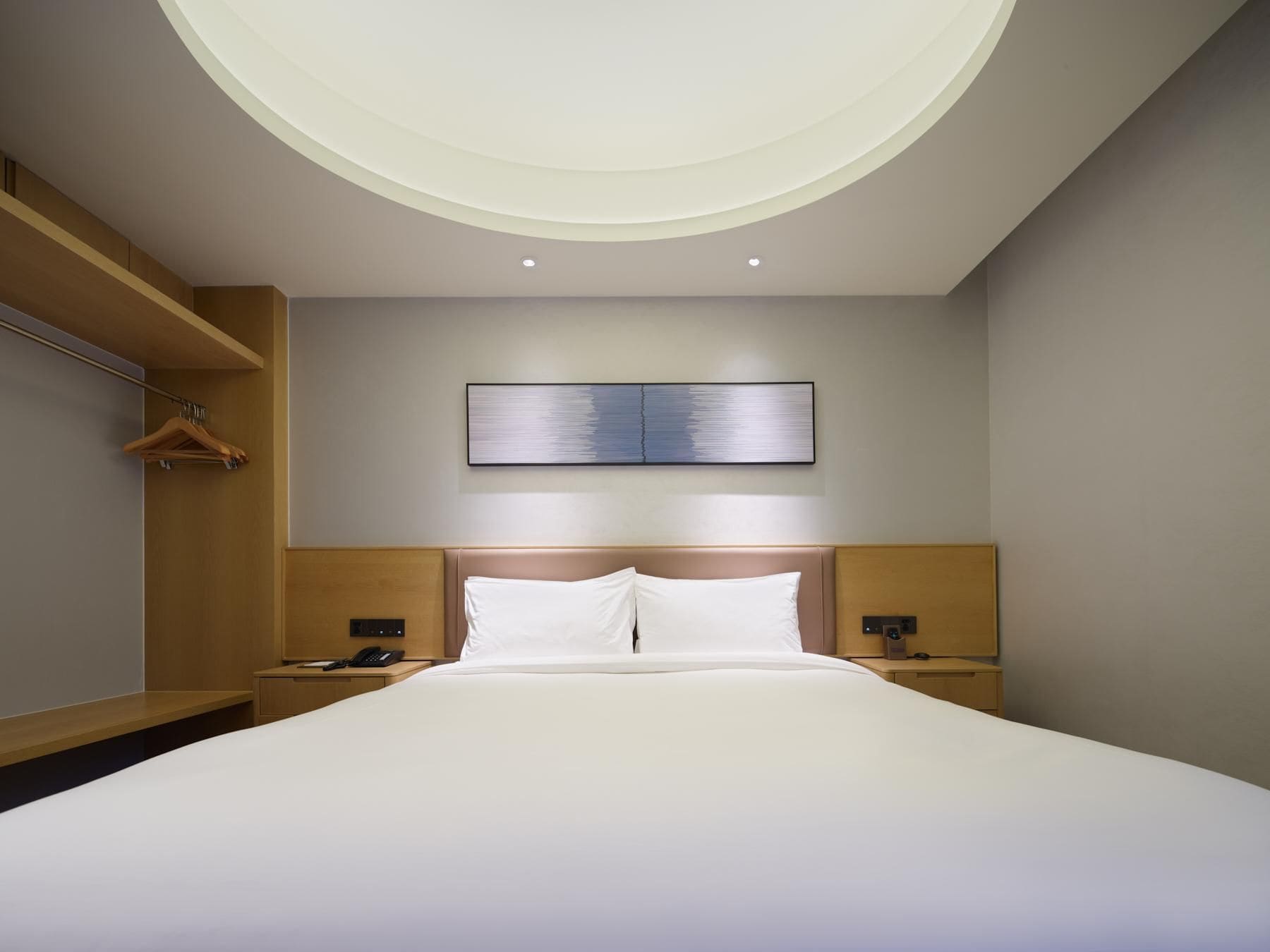 Quarto de hotel moderno com uma cama grande, cabeceira de madeira, mesas de cabeceira, uma obra de arte na parede e um claraboia circular.
