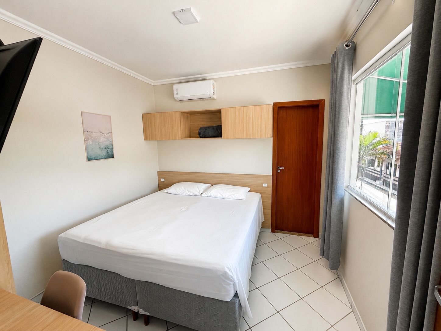 Quarto de hotel com cama, escrivaninha, janela com cortinas, porta e unidade de ar condicionado.